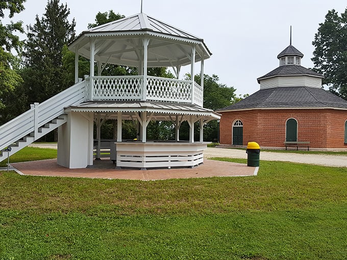 A classic white gazebo stands ready for summer concerts while a brick structure waits nearby. Small-town gathering spots that bring communities together through music and celebration.
