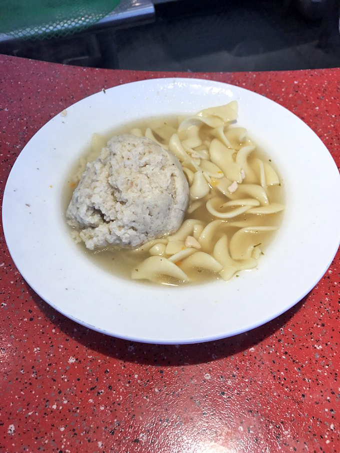 Behold the star attraction: a matzo ball floating majestically in golden broth with egg noodles, like comfort in a bowl waiting to cure whatever ails you.