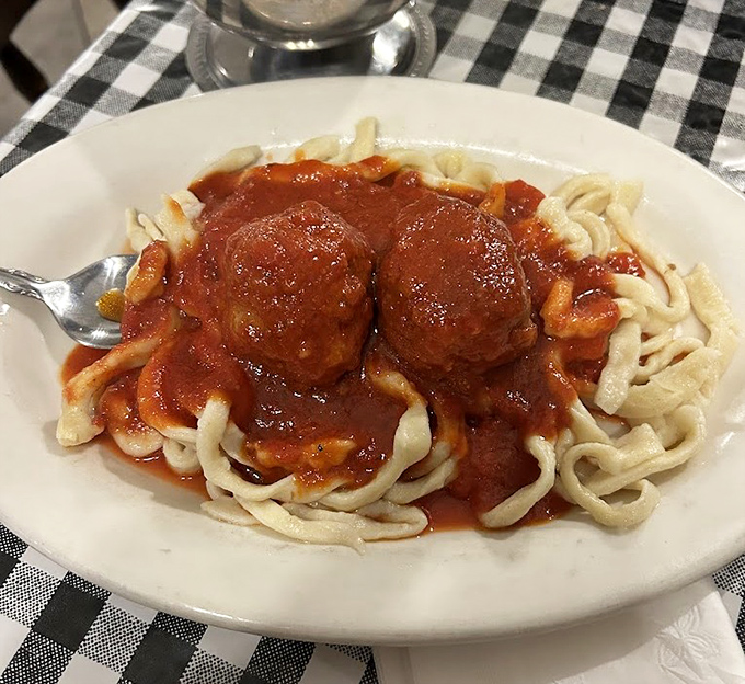 Behold the stars of our show: two perfectly formed meatballs resting atop homemade pasta, swimming in a sauce that whispers secrets of generations past.