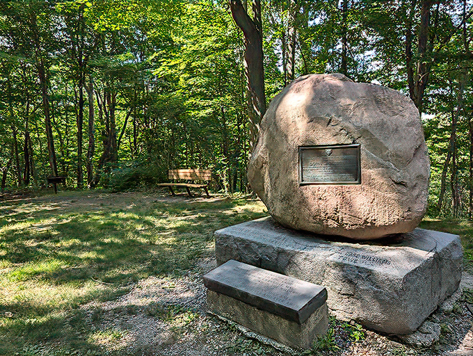 A memorial stone stands sentinel in the dappled sunlight, telling stories of the past while forest benches invite contemplation.