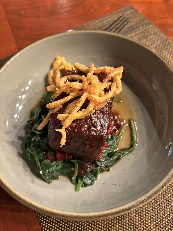 The braised short rib sits like a king atop its throne of wilted greens, crowned with crispy onions that shatter at first bite.