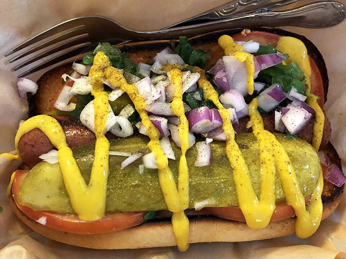 A Chicago-style hot dog that's dressed to impress, with vibrant yellow mustard zigzagging across a landscape of crisp pickles and fresh toppings.