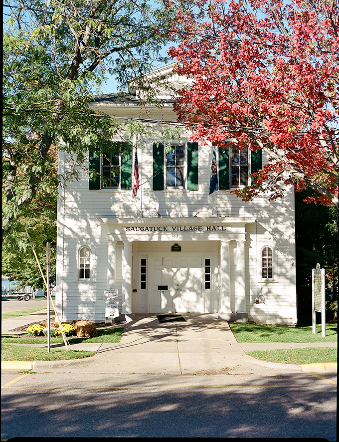 Saugatuck Village Hall stands proudly amid autumn splendor, its crisp white facade and green shutters embodying small-town charm that Norman Rockwell would have rushed to paint.