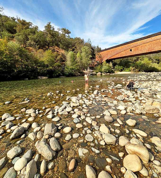 Smooth river stones create nature's mosaic beneath crystal-clear waters, inviting summer visitors to cool their toes while admiring the bridge's elegant silhouette.