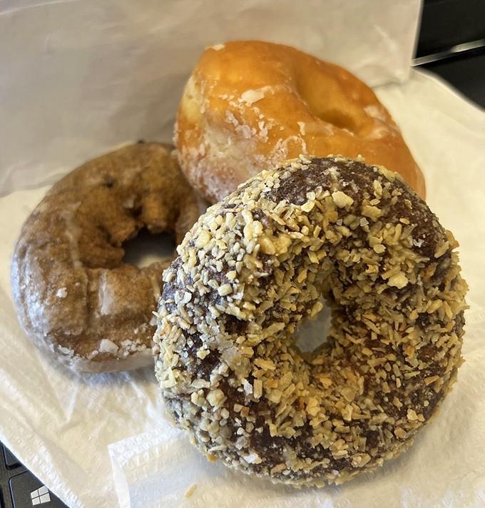 Three donuts representing the holy trinity of breakfast pastries: glazed perfection, nutty crunch, and that mysterious middle child nobody regrets trying.