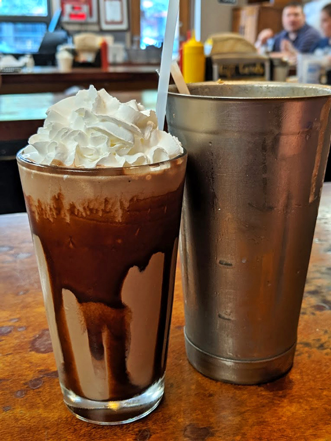 The chocolate shake arrives with whipped cream mountain and a side of nostalgia in that classic metal mixing cup—proof that some traditions need no improvement.
