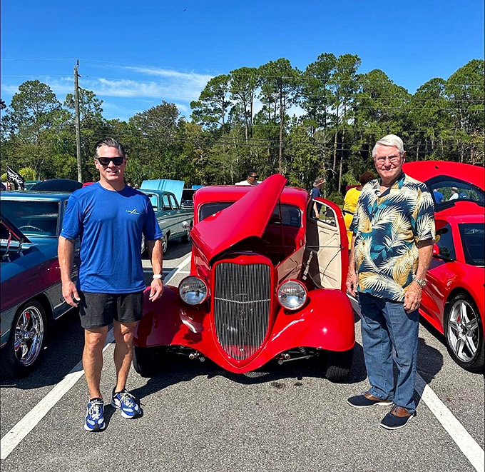 Car enthusiasts sharing stories beside a cherry-red hot rod. The universal language of automotive passion needs no translation on a sunny Florida day.