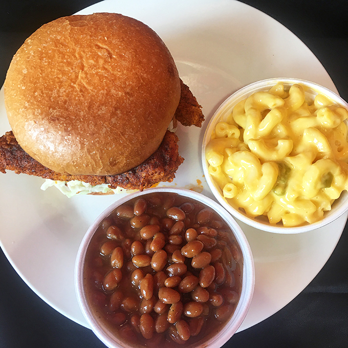 A hot chicken sandwich holds court on a white plate, flanked by mac and cheese and baked beans—the holy trinity of Southern comfort.