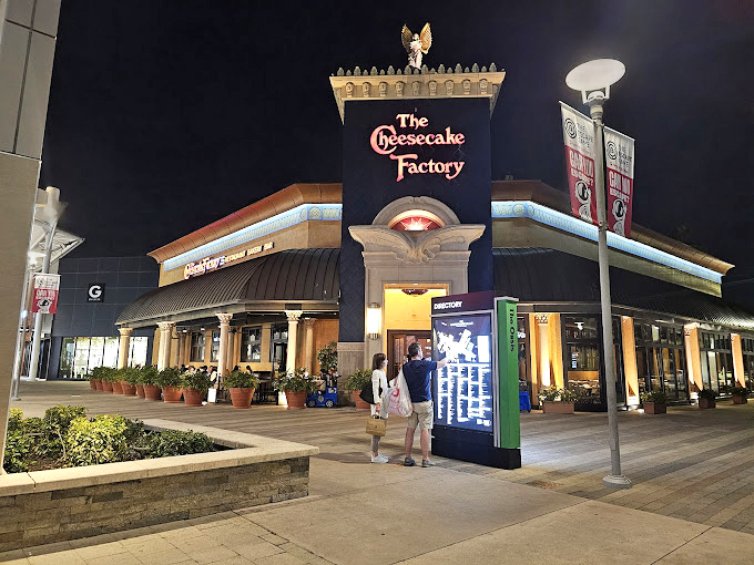 The Cheesecake Factory glows invitingly at dusk, promising a delicious reward for shoppers who've earned their calories after hours of retail cardio.