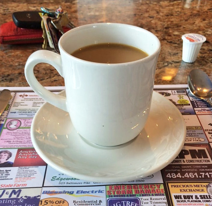 The humble white coffee mug&mdash;diner civilization's most perfect vessel, delivering liquid motivation against a backdrop of local newspaper ads.