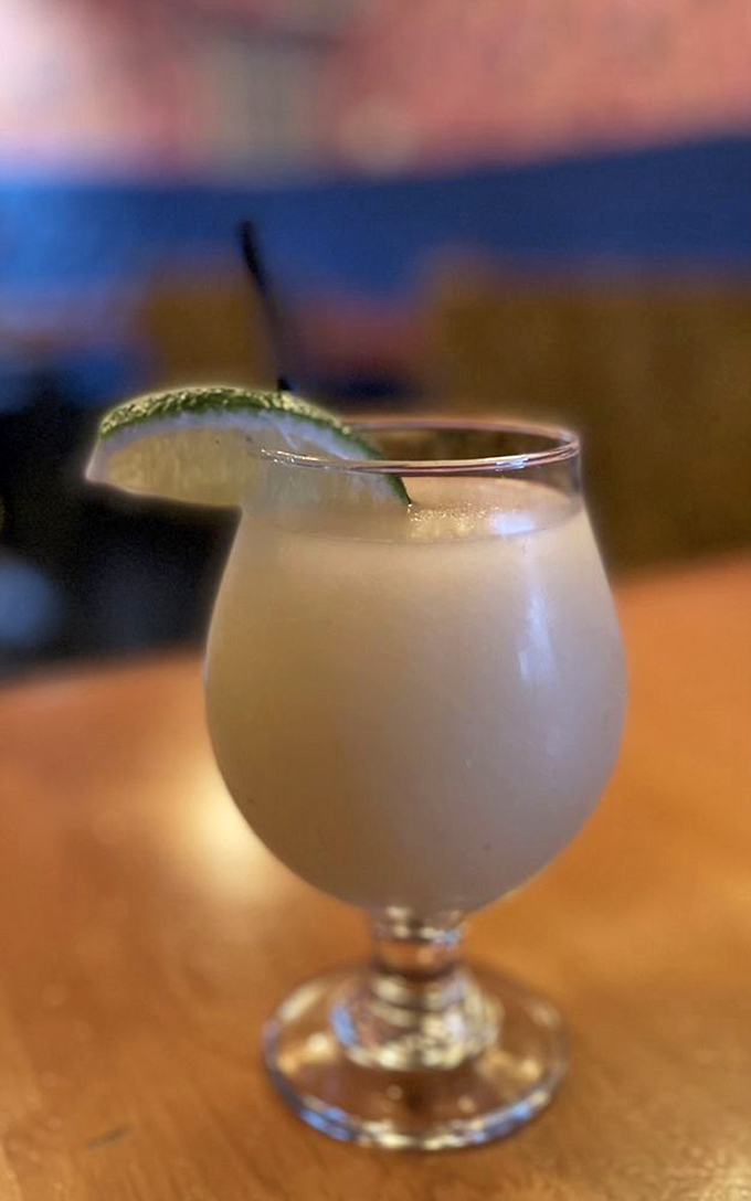 A frosty margarita with a lime garnish sits ready to refresh. In the blurred background, the restaurant's signature blue and pink decor creates the perfect California mood.
