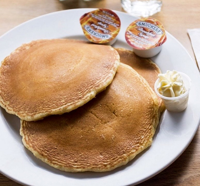 Pancakes so perfectly golden they belong in Fort Knox. These fluffy discs of joy arrive with butter melting seductively and tiny jam cups standing by for duty.