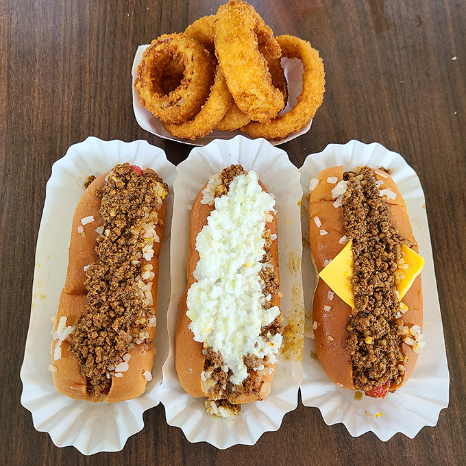The holy trinity of hot dog heaven: chili, slaw, and classic&mdash;with onion rings standing guard like crispy sentinels of satisfaction.