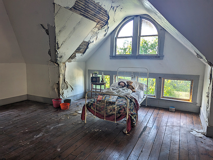 Sunlight and shadows play across weathered floorboards in this attic bedroom, where a solitary iron bed frame stands as a silent witness.