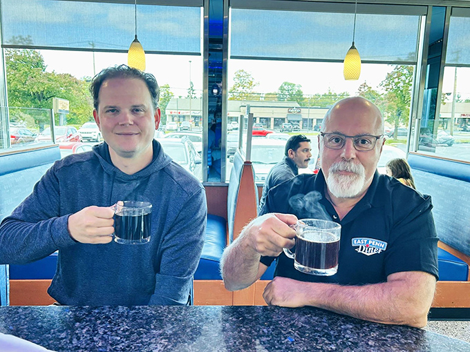 Coffee mugs raised in a toast to good conversation. The blue booths behind them have witnessed countless local stories unfold.
