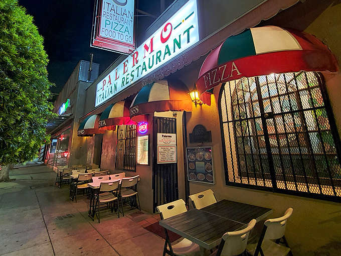 By night, Palermo transforms into a glowing oasis of Italian comfort, its illuminated sign and colorful umbrellas beckoning hungry souls like moths to a carb-laden flame.