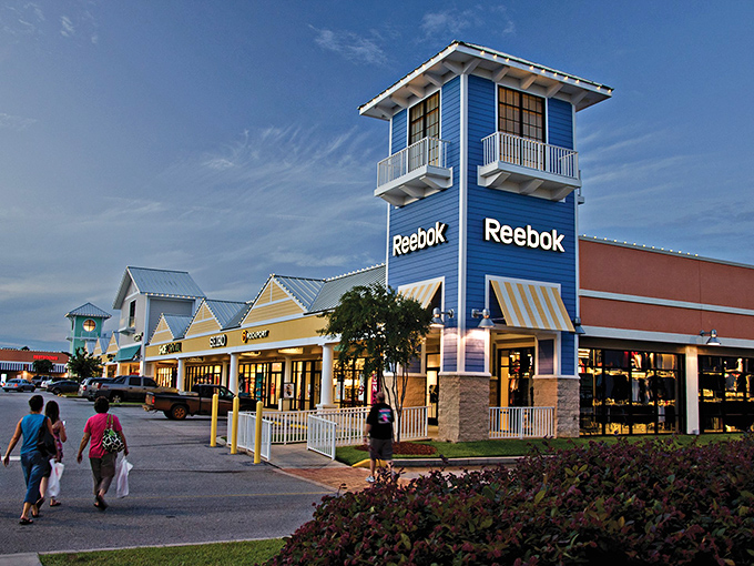 The blue Reebok tower stands like a retail lighthouse, guiding bargain hunters to shores of savings while palm-lined walkways invite exploration.