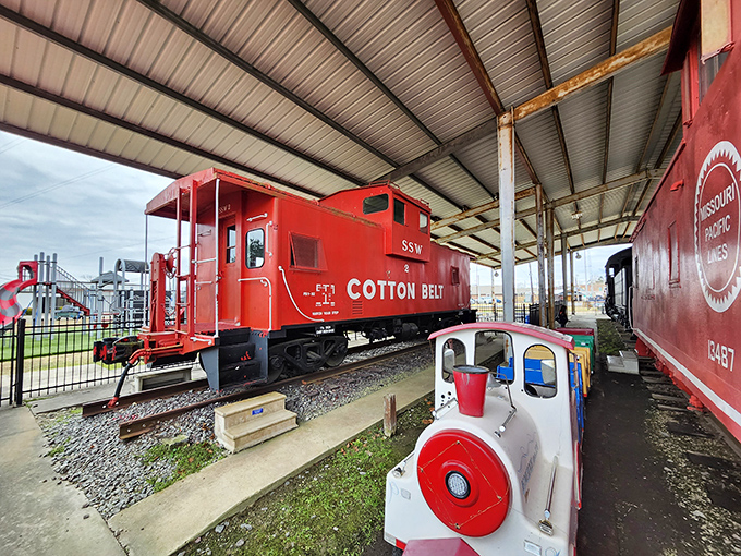 The iconic red Cotton Belt caboose stands proudly under shelter, a crimson reminder of railroading's golden age. The kid-friendly train ride in the foreground adds a playful touch for young engineers-in-training.