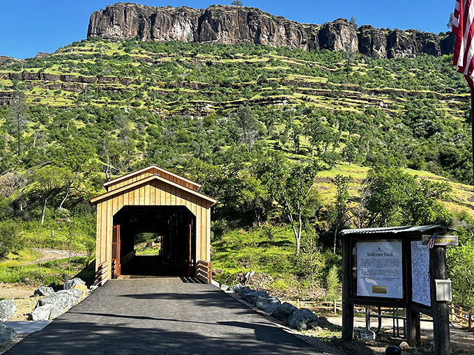 The honey-colored covered bridge sits at the foot of dramatic rocky cliffs, its warm wooden frame glowing under the bright California sun as it invites visitors into the landscape beyond.