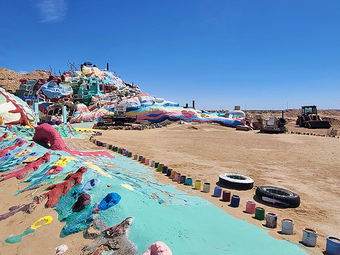 A technicolor dream rises from the desert floor, where paint cans stand guard like tiny sentinels protecting this vibrant masterpiece.