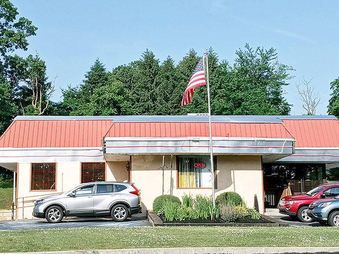 The American flag stands tall above this unassuming roadside gem, where culinary magic happens behind that humble beige facade and distinctive red roof.