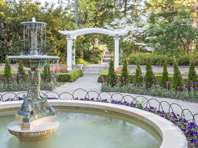 A tiered fountain stands sentinel among purple blooms, like nature's concierge welcoming you to this garden paradise. The white pergola beckons you forward.