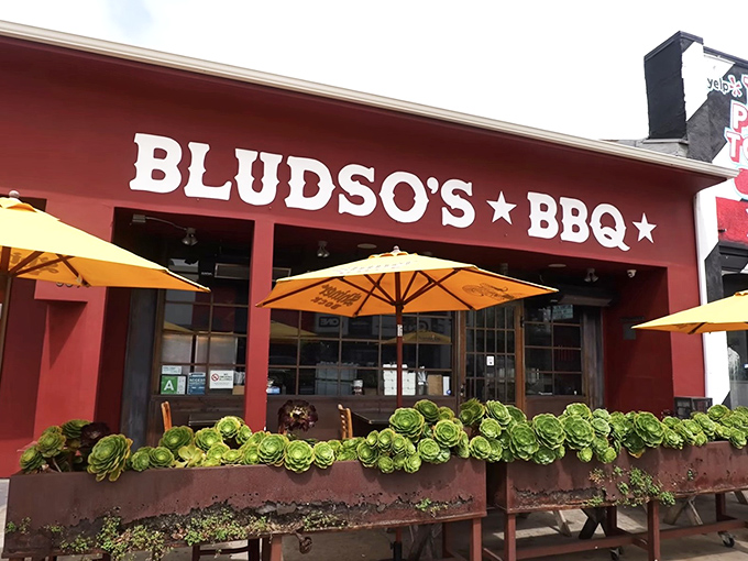 The crimson facade of Bludso's BBQ stands out like a beacon of hope for the hungry, promising smoky salvation under those cheerful yellow umbrellas.