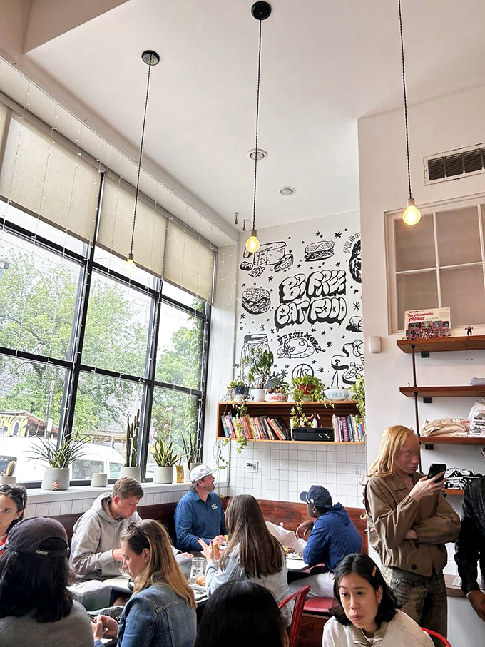 Sunlight streams through tall windows while diners enjoy their sandwiches beneath a playful food-themed mural.