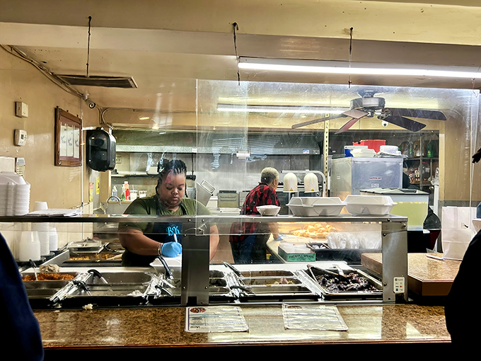 Behind the glass, culinary magic happens with practiced hands moving efficiently between bubbling pots of Southern goodness.