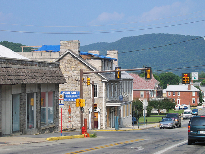 Bright summer sun highlights a street corner lined with historic stone and clapboard buildings, leading the eye toward the rolling green hills in the distance under a clear blue sky.