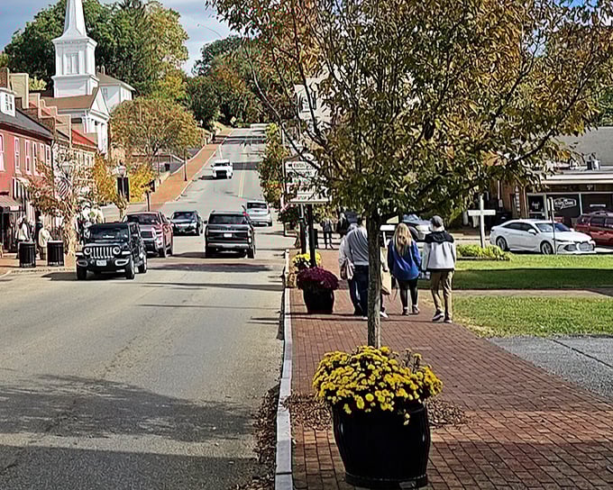 Fall colors frame Main Street as pedestrians stroll past planters bursting with chrysanthemums. The white church steeple stands sentinel over this postcard-perfect scene.