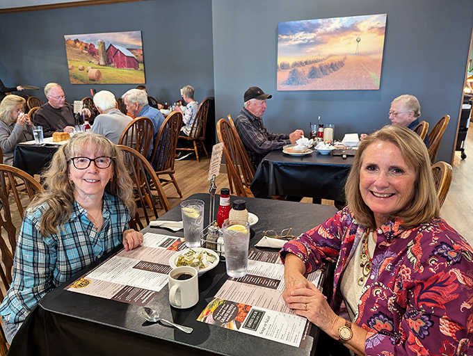 Happy diners sharing stories over steaming plates—the universal language of good food bringing strangers together under rustic farm paintings.