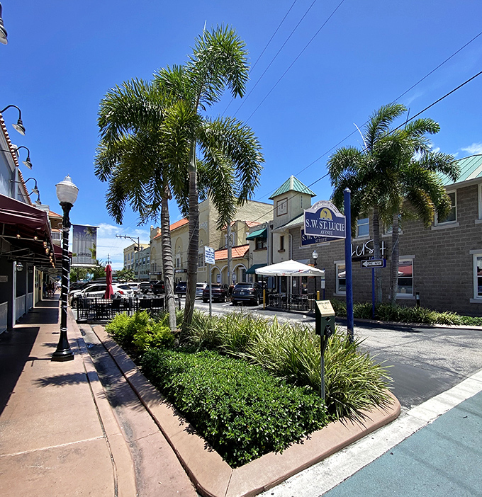 Sunlight pours down on a tropical downtown street, where feathery palm trees and lush green planters line the sidewalk beside historic buildings under a vibrant blue sky.