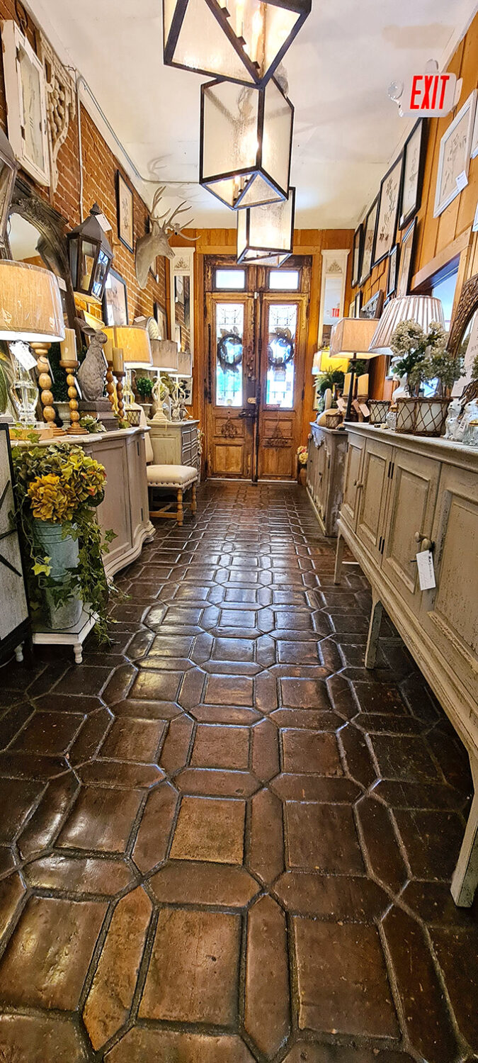 Terracotta tiles lead down a hallway of farmhouse chic, where pendant lights cast a warm glow over distressed cabinets and botanical arrangements.