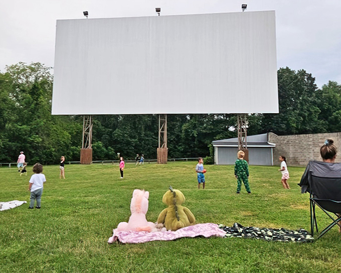 Between features, kids reclaim the grassy stage beneath the towering screen, burning energy while stuffed animals patiently guard blanket real estate.