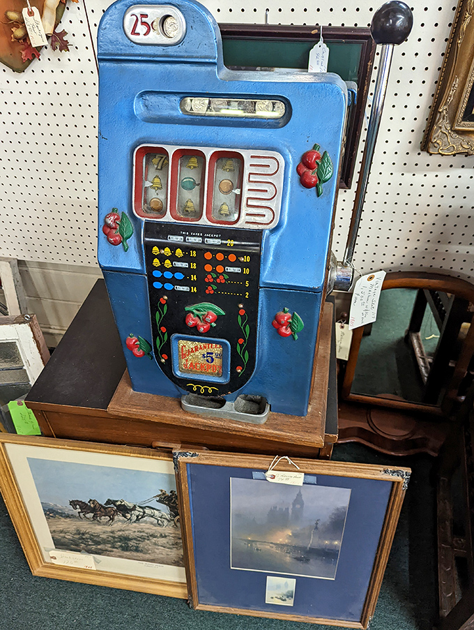 Cherry-decorated slot machine stands guard over framed landscapes, proving that gambling and art make surprisingly compatible roommates in antique mall logic.