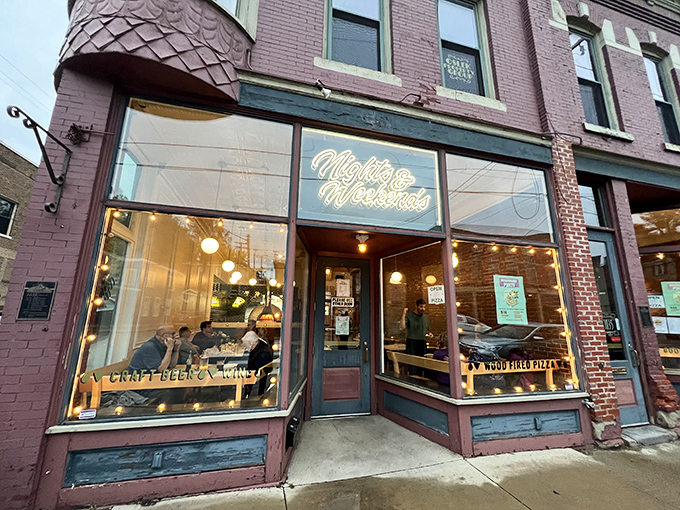 Nights & Weekends Pizza glows with inviting warmth behind vintage storefront windows. The neon sign promises comfort food in a space where conversations flow as freely as the drinks.
