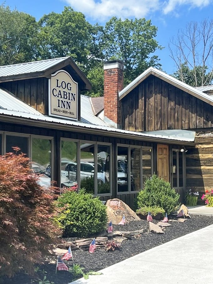 Summer sunshine highlights the Log Cabin Inn's authentic wooden exterior and meticulous landscaping. Small American flags add patriotic charm to this culinary destination.