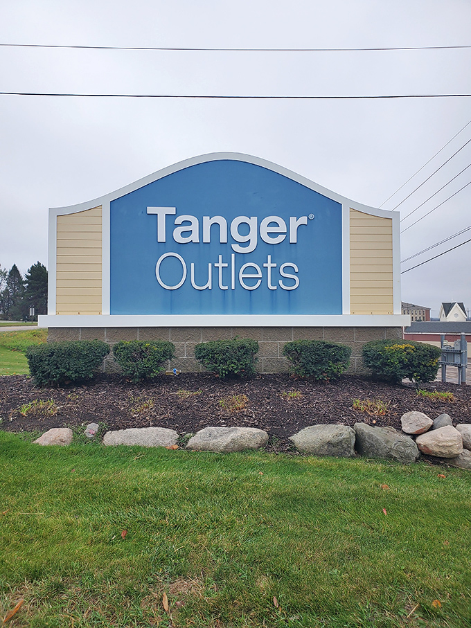 The blue and beige Tanger Outlets sign stands as a retail lighthouse, guiding bargain-seeking ships safely to harbor through seas of full-priced temptation.