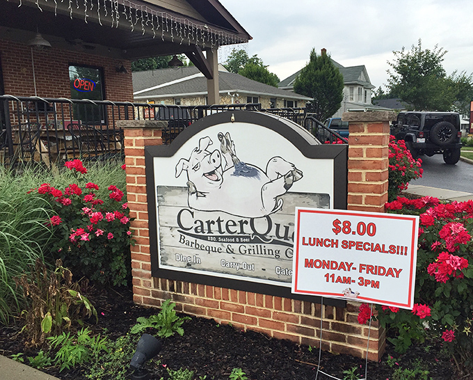 The welcoming sign at CarterQue, surrounded by vibrant flowers, promises BBQ bliss with that charming lunch special that's practically begging you to visit.