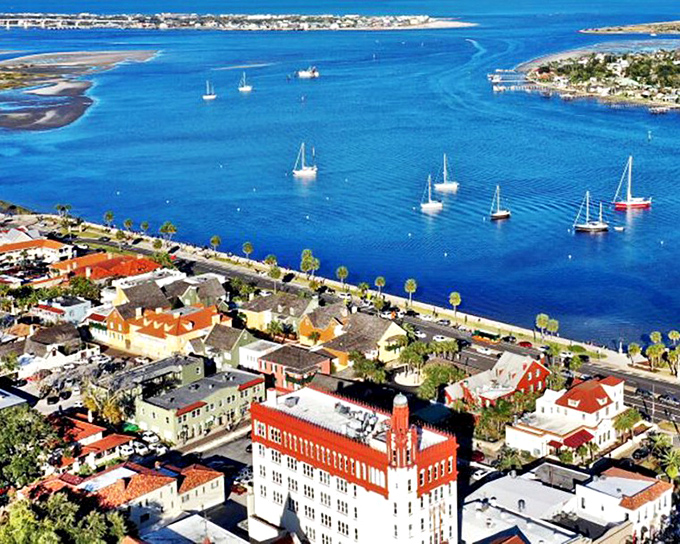 That stunning red-roofed buildings anchors the waterfront like a beacon, with sailboats bobbing peacefully in impossibly blue water below.