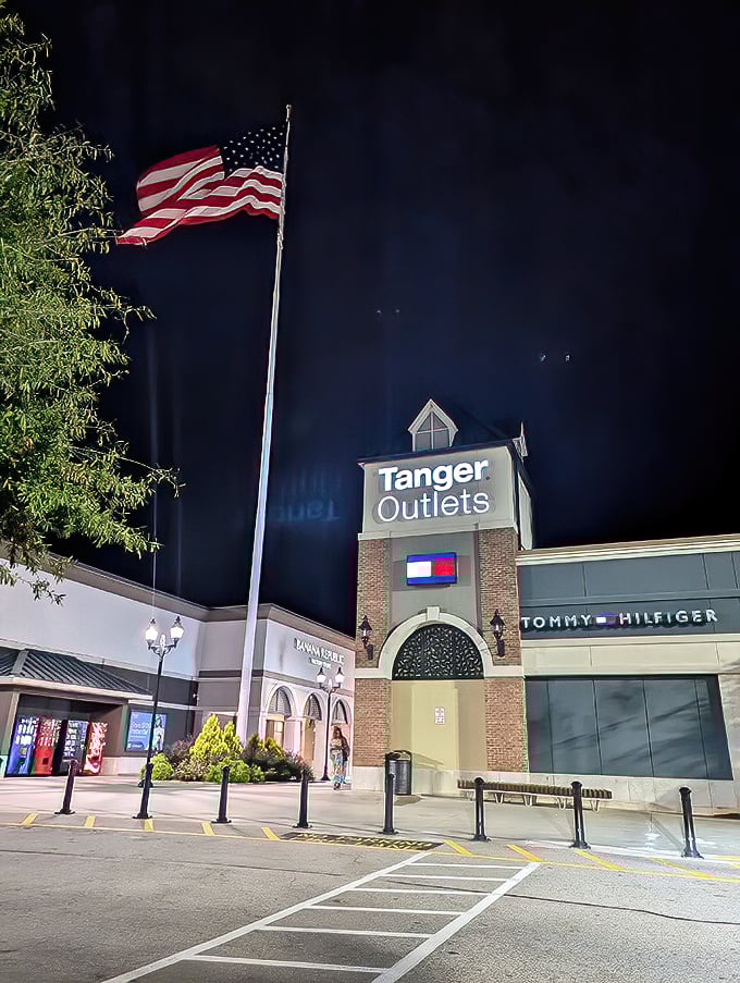 Night shopping takes on a patriotic glow at Tanger's entrance, where Old Glory waves proudly above designer discounts. The American dream: life, liberty, and the pursuit of bargains.
