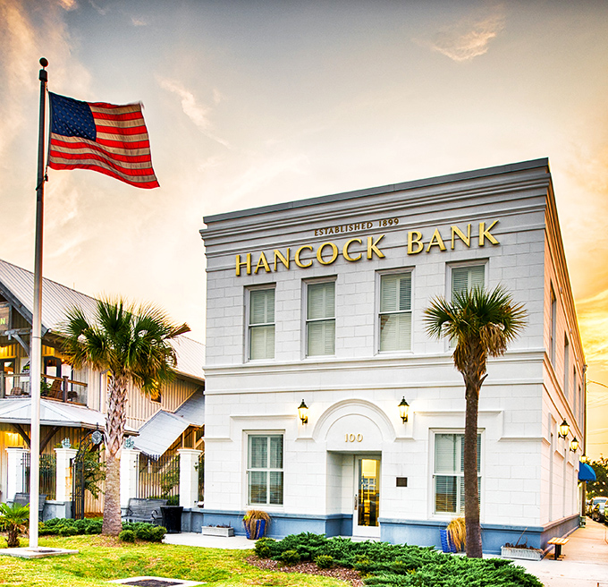 Hancock Bank stands as a gleaming sentinel of Southern prosperity, where palm trees and patriotism frame a building that's weathered more storms than your aunt's holiday dinners.
