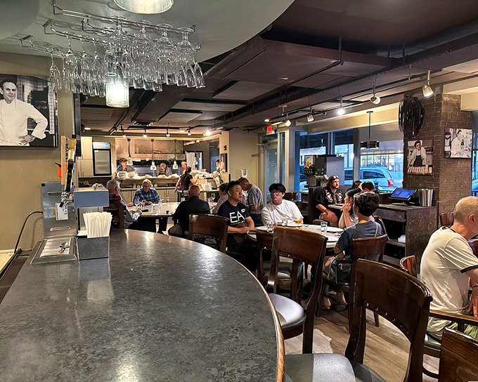 The bustling interior blends rustic charm with urban sophistication, where wine glasses dangle overhead like crystal stalactites in a cave of culinary wonders.