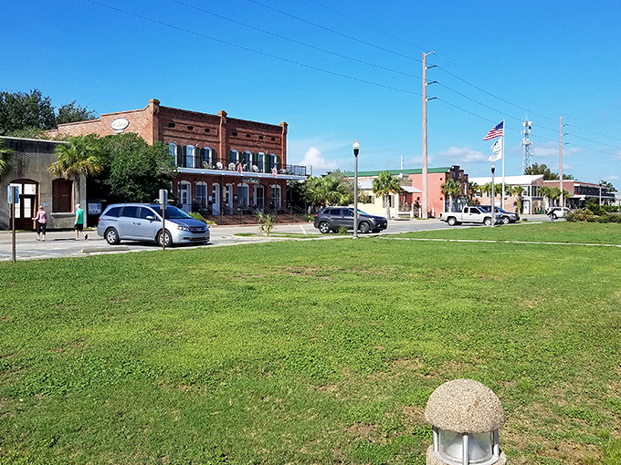 Warm sunlight streams across a wide green park, overlooking a street lined with historic brick buildings and modern storefronts under a clear blue sky.