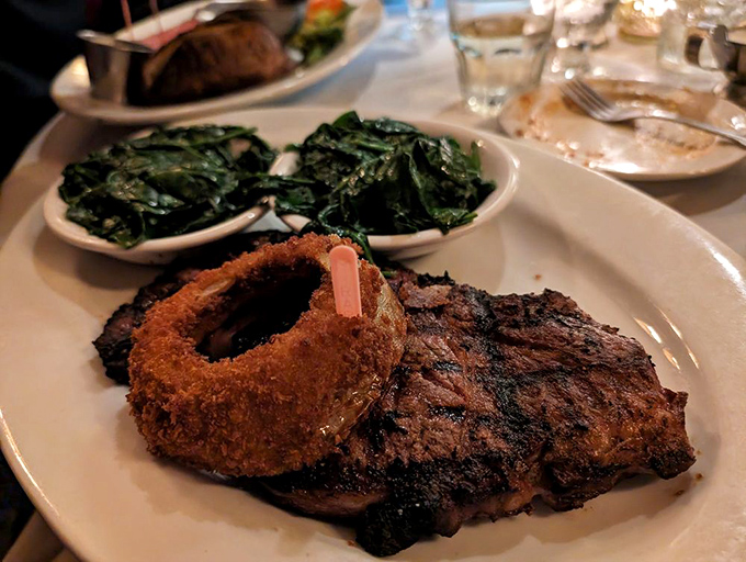 Behold the star of the show: a perfectly seared ribeye alongside crispy onion rings and saut&eacute;ed greens. Meat poetry on a plate.