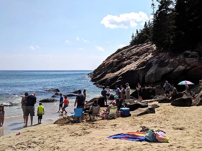 Beach day sociology in action: families claiming their sandy real estate beneath the watchful eye of ancient granite. The timeless dance of summer unfolds here daily.