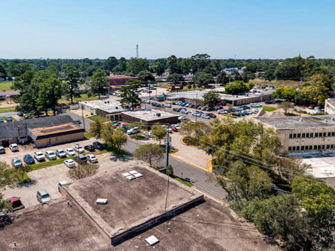 Clear midday sun shines over a flat-roofed commercial center, surrounded by dense green trees and multiple busy parking lots in this suburban aerial view.