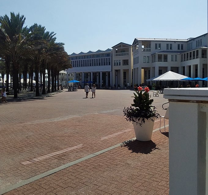 Bright midday sun shines over a spacious brick plaza, where uniform white buildings and a grove of tall palm trees create a distinct resort town aesthetic.