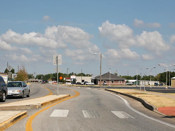 Fluffy white clouds float across the bright blue sky over a curving suburban road, leading past low commercial buildings and a row of streetlights on a sunny afternoon.